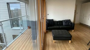 Uma sala de estar moderna com um sofá e um banquinho pretos, piso de madeira e uma grande janela com vista para um edifício.