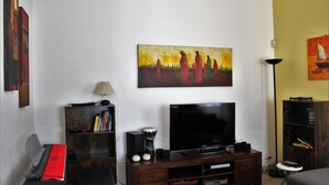 Uma sala de estar com televisão de tela plana, estante de livros, luminária e um quadro na parede.
