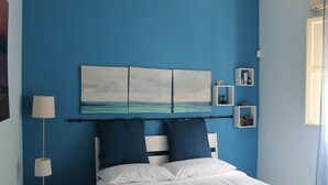 Quarto com cama, mesa de cabeceira, luminária, pintura e prateleira com objetos.