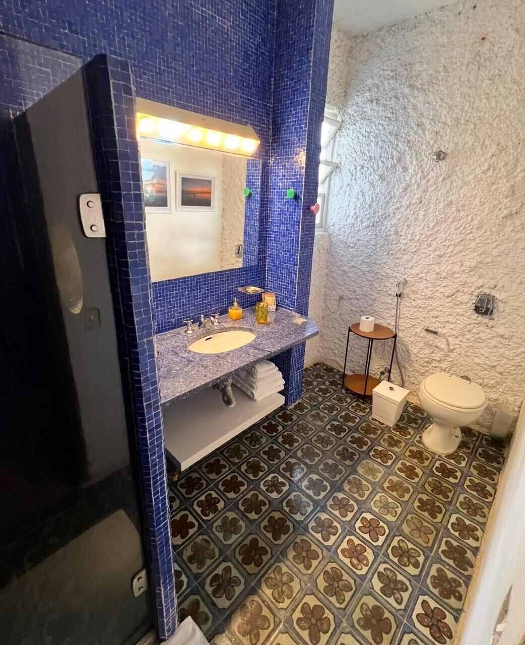 Um banheiro com parede revestida em azulejos azuis, pia com espelho, vaso sanitário e piso estampado.