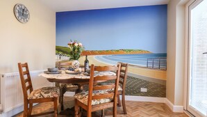 Uma sala de jantar com um mural de praia, cadeiras de madeira, uma mesa posta com pratos, um vaso de flores e um relógio na parede.
