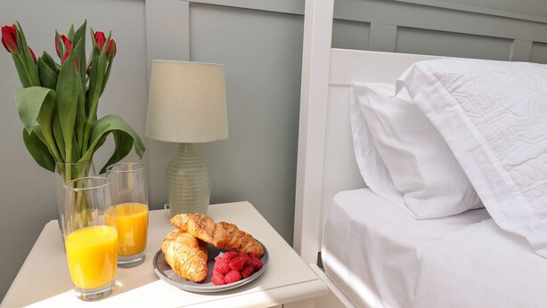Uma cama bem arrumada com lençóis brancos, uma mesa de cabeceira com abajur, um prato com croissants e morangos, e um copo de suco de laranja.
