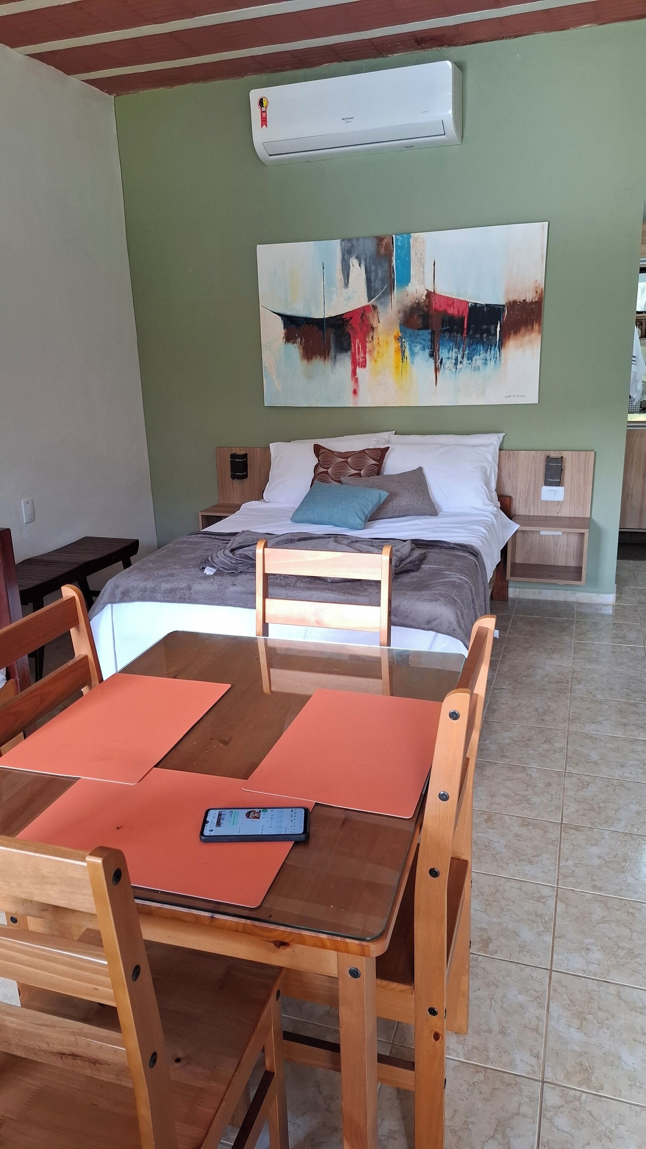 Um quarto com uma cama, um quadro, uma mesa com cadeiras e um ar condicionado.