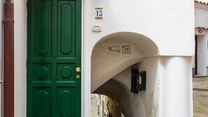Uma porta verde com uma pequena janela acima, uma parede branca com uma placa de número e um arco de pedra que dá para um corredor.