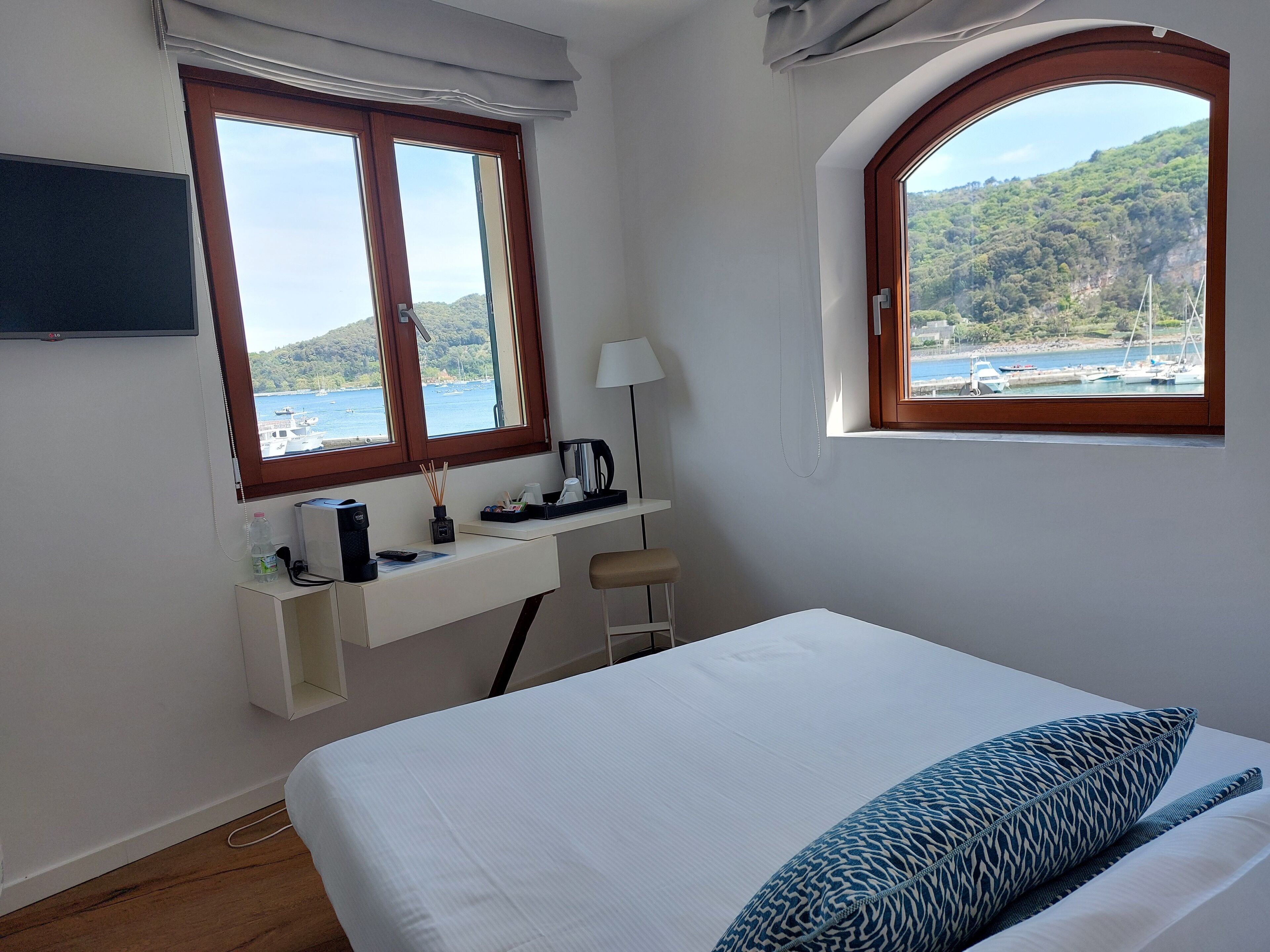 Quarto de hotel com cama, escrivaninha com luminária, televisão e vista para o mar e colinas.