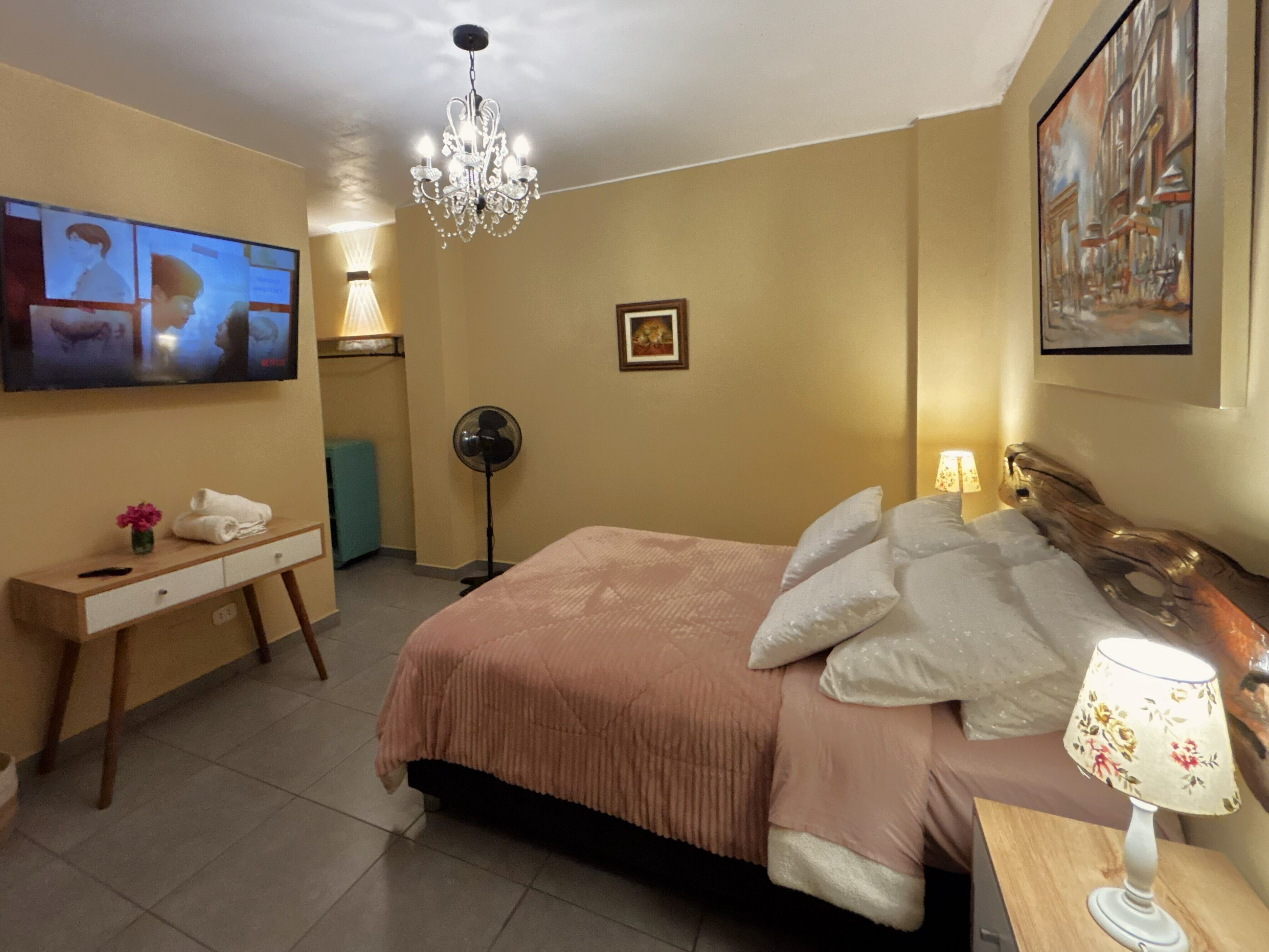 Quarto de hotel com cama, mesa de cabeceira, abajur, televisão e um ventilador.