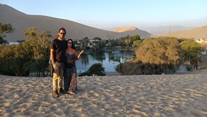 Um casal está em uma praia de areia, com um lago e montanhas ao fundo.