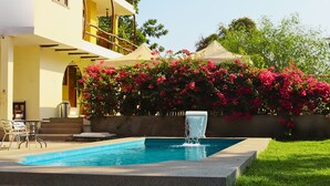 Uma piscina com um elemento aquático, um edifício de dois andares com varandas, e exuberantes plantas de bougainvillea cor-de-rosa.