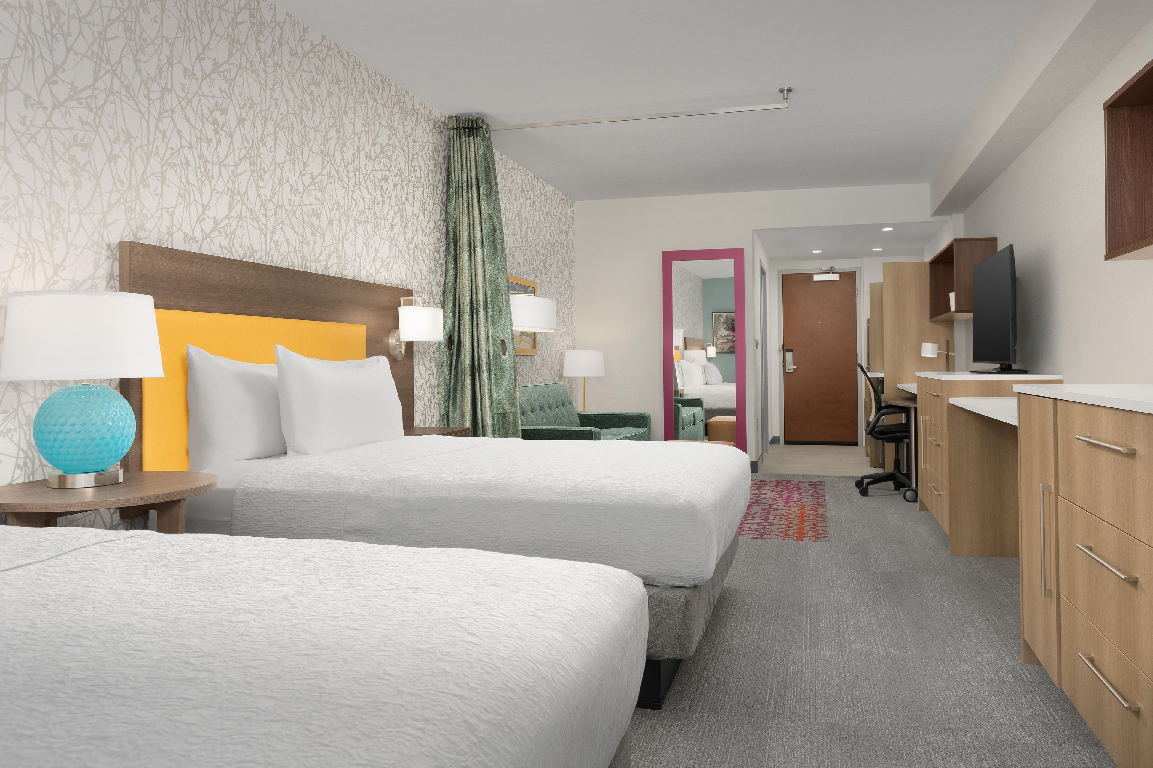 Quarto de hotel com duas camas, uma escrivaninha, televisão e banheiro visível por uma porta aberta.