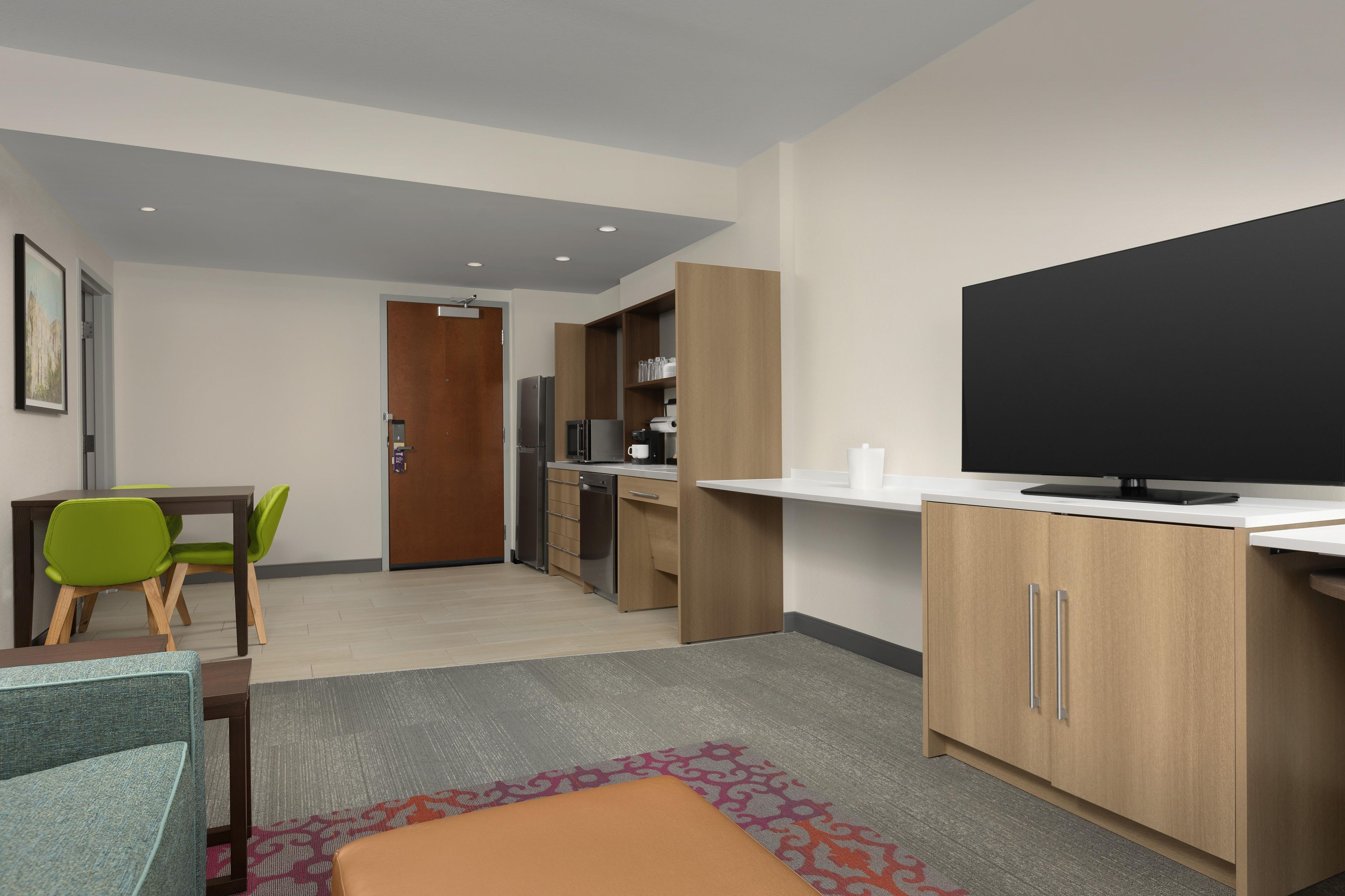 Quarto de hotel moderno com televisão de tela plana, armários de madeira, área de refeições com mesa e cadeiras e cozinha equipada.