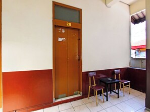 Quarto com porta marrom identificada com o número 211, uma pequena mesa com duas cadeiras e uma janela com toldo vermelho.