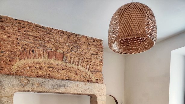 Quarto com cabeceira de pedra, cama com várias almofadas, mesa de cabeceira com espelho e um prato, e luminária suspensa.