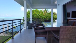 Varanda moderna ao ar livre com mesa de vidro e cadeiras de vime, com vista para um mar azul límpido e vegetação exuberante.