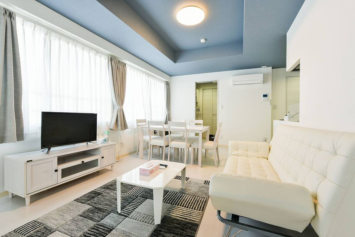 Uma sala de estar moderna com televisão de tela plana, mobiliário branco e uma grande janela com cortinas.
