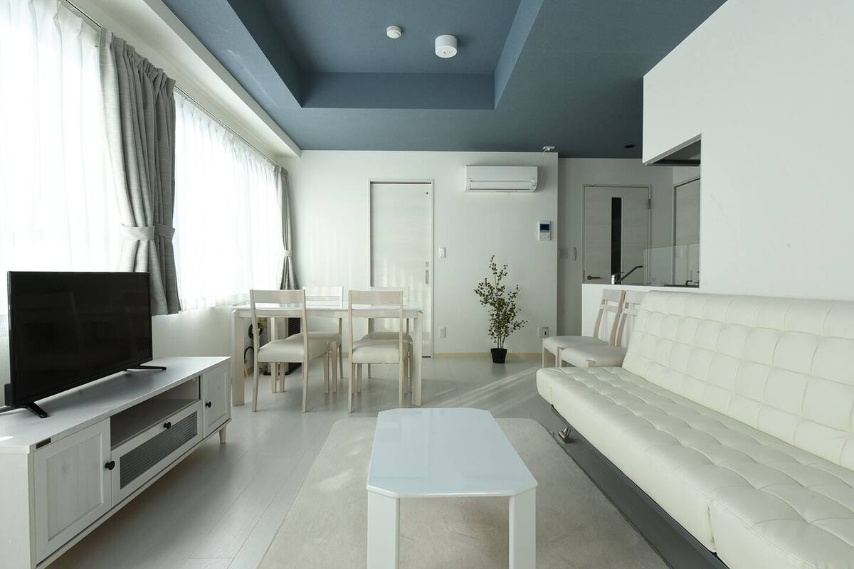 Uma sala de estar moderna com televisão de tela plana, móveis brancos, uma luminária de teto e um vaso com planta.