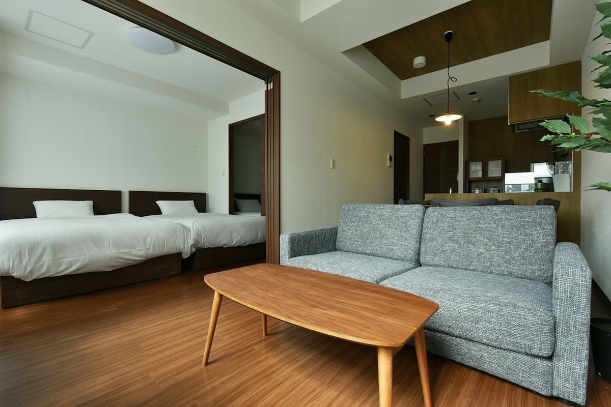 Quarto de hotel moderno com chão de madeira, sofá cinza, duas camas de solteiro, mesa de centro de madeira e cozinha visível através de uma porta aberta.