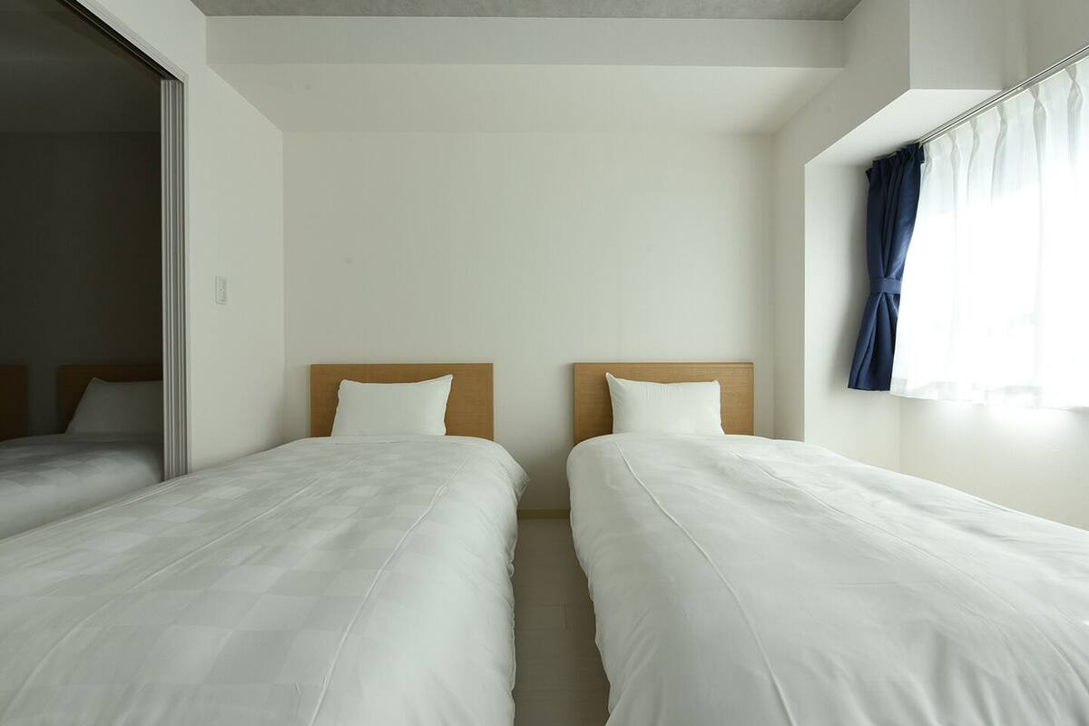 Quarto de hotel com duas camas de solteiro, cabeceira de madeira, janela com cortinas leves e porta que dá para outro cômodo.