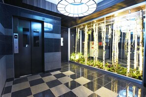 Um elevador com mostrador digital e botões de emergência, piso xadrez e parede de vidro com jardins verticais.