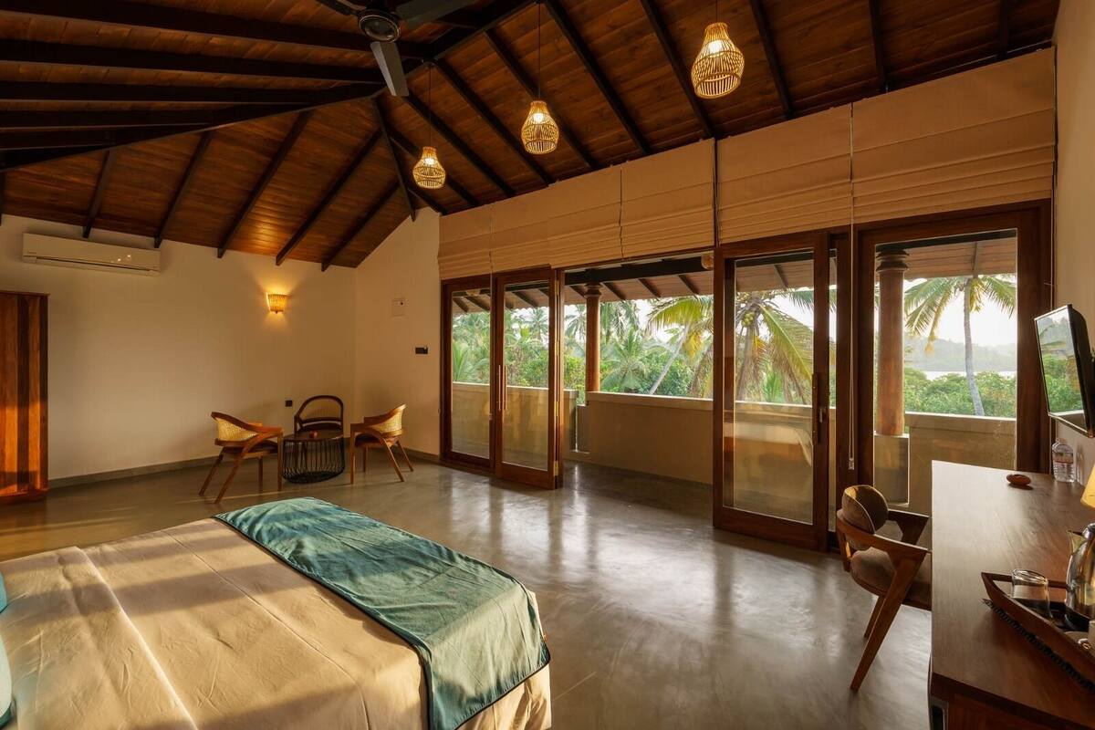 Quarto amplo com cama, escrivaninha e cadeiras. Janelas grandes com vista para palmeiras e uma varanda.