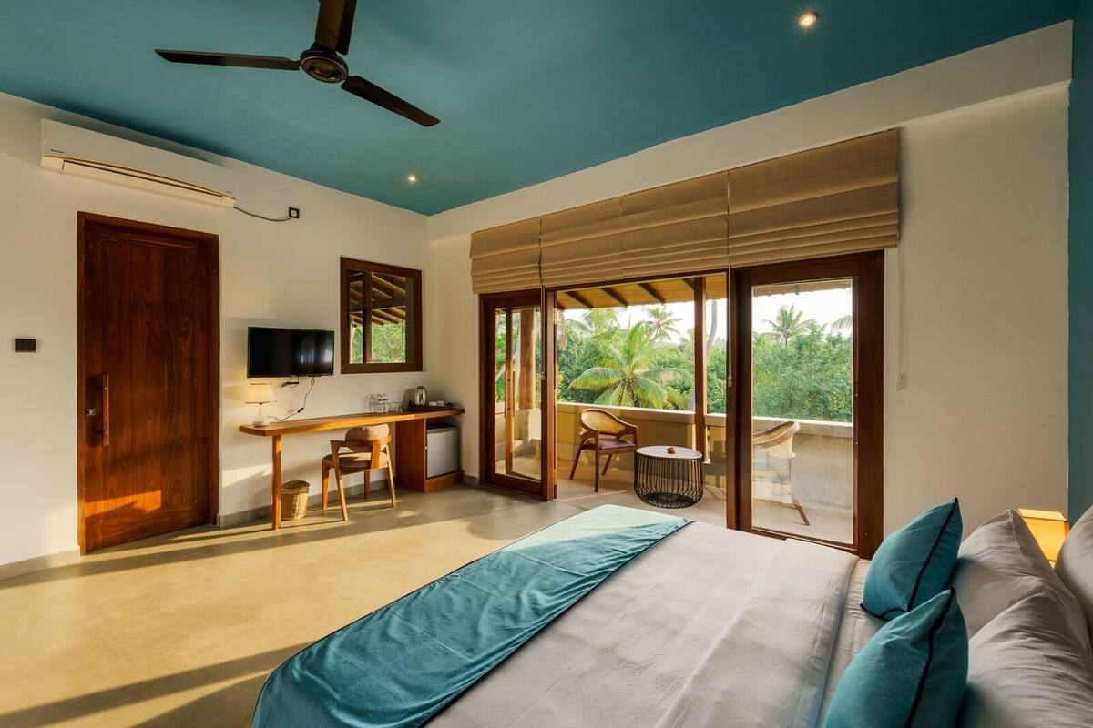Quarto com cama, escrivaninha, televisão e varanda com vista para palmeiras.
