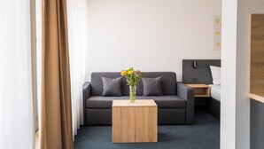 Quarto de hotel moderno com um sofá cinzento, uma mesa de centro de madeira e uma pequena mesa de cabeceira com uma luminária.