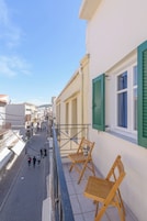 Uma rua estreita com edifícios de ambos os lados, uma varanda com cadeiras de madeira e um céu azul claro.