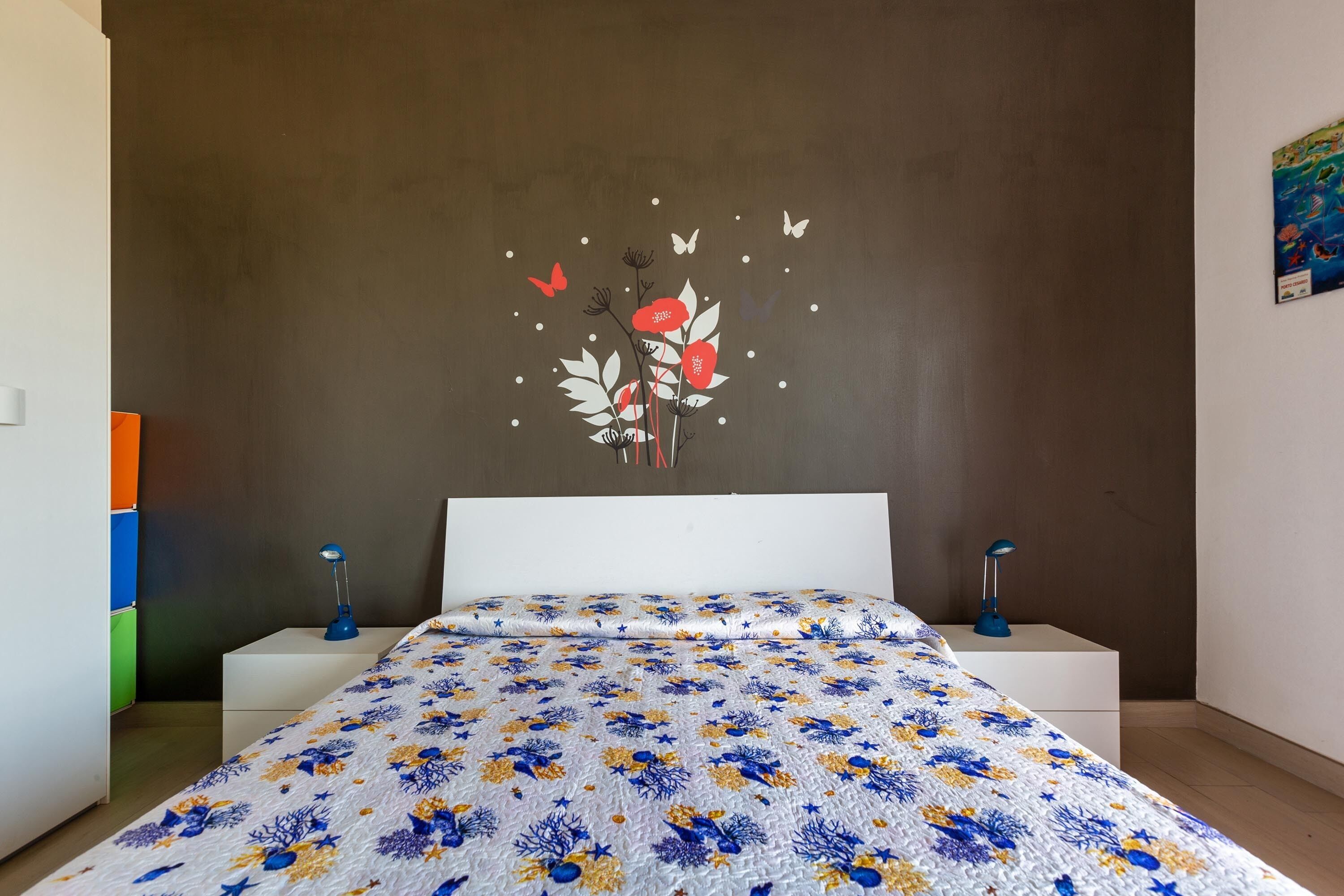 Quarto com cama, duas mesas de cabeceira, um mural na parede e vista parcial de um guarda-roupa.