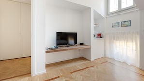 Quarto moderno e minimalista com piso de madeira, uma TV de tela plana em um suporte de madeira, uma mesinha com um controle remoto e uma janela com cortinas leves.