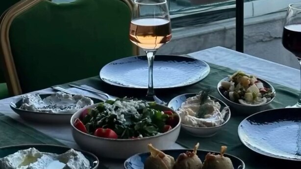 Mesa posta com diversos pratos e um copo de vinho, com vista para paisagem urbana e um corpo de água, além de edifícios.