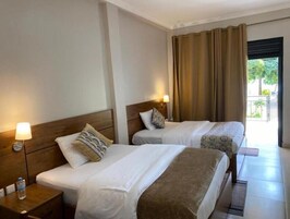 Quarto de hotel com duas camas, uma mesa de cabeceira com abajur, uma janela com cortinas e vista para o verde lá fora.