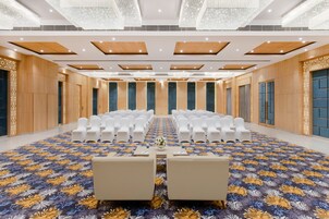 Uma sala de conferências ampla, com fileiras de cadeiras brancas, uma mesa central decorada com flores e um tapete estampado.