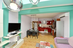 Uma sala de estar moderna com televisão de tela plana, mesa de jantar e cadeiras. Há uma área de cozinha com armários vermelhos e uma geladeira.