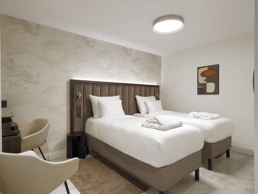 Quarto de hotel moderno com uma cama grande, duas mesas de cabeceira, uma cadeira e uma pequena escrivaninha.