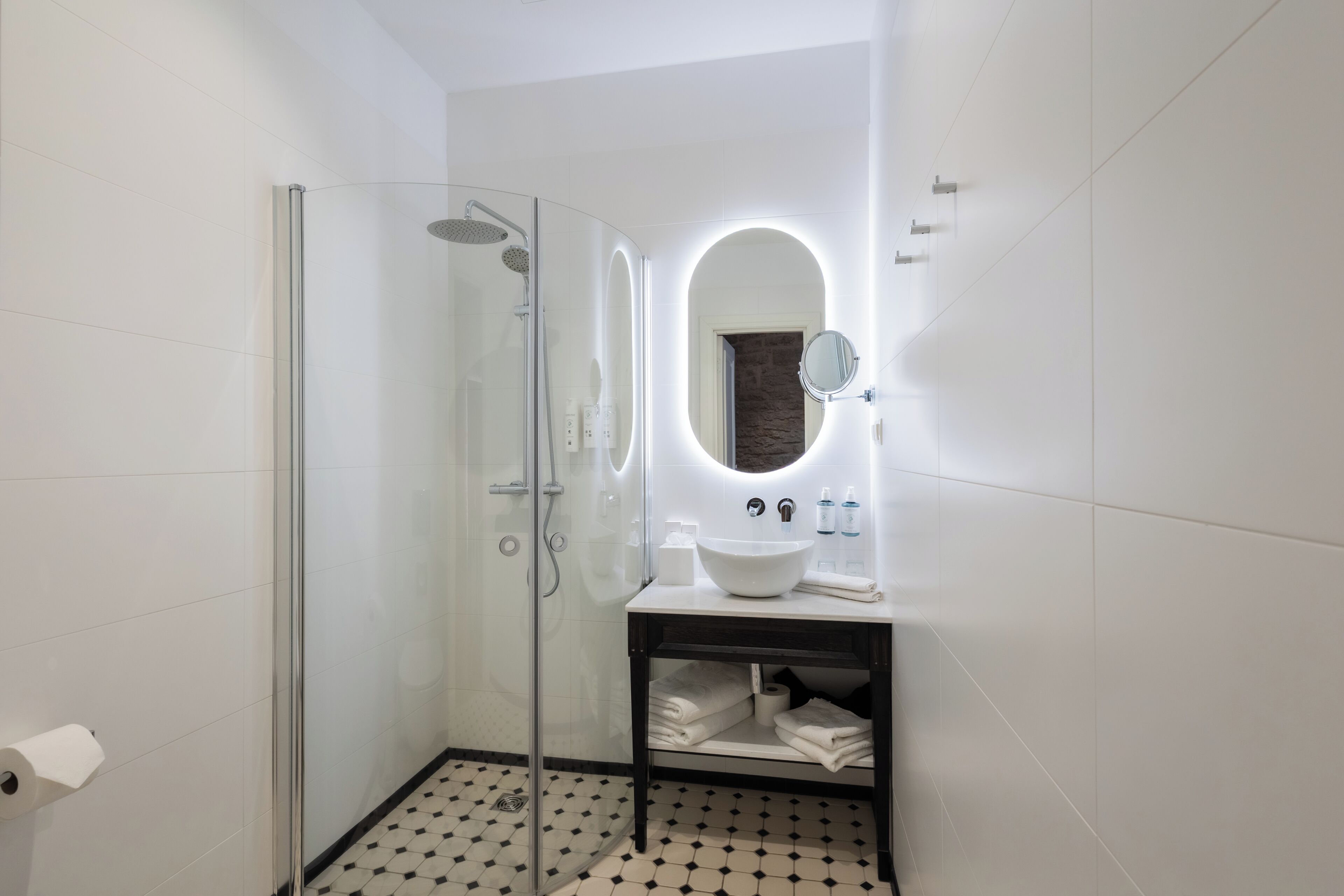 Uma casa de banho moderna com cabine de duche em vidro, lavatório branco, espelho redondo e chão em mosaico preto e branco.
