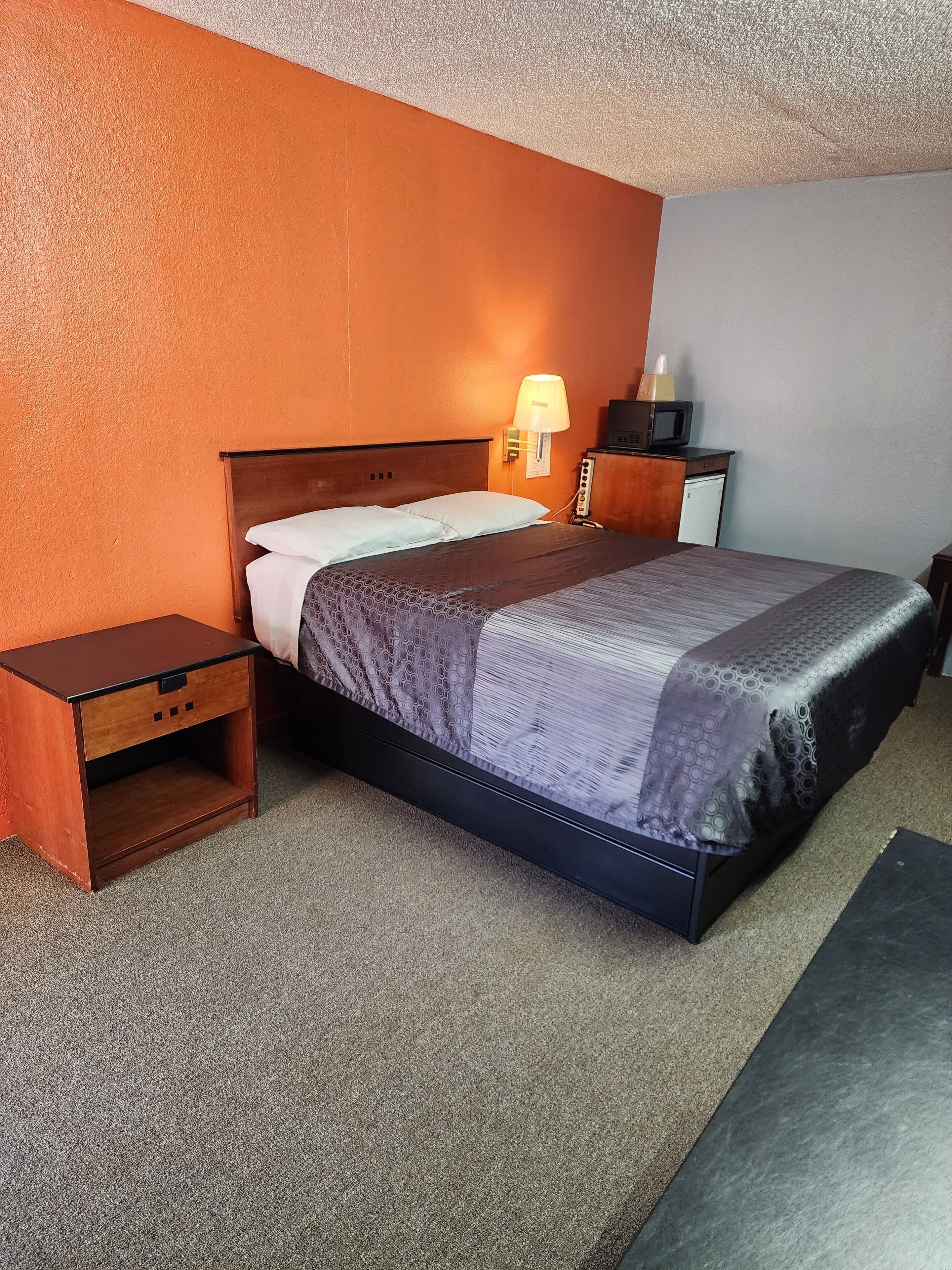 Quarto de hotel com uma cama, mesa de cabeceira, abajur, micro-ondas e um pequeno armário.