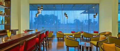 Uma área de bar com um balcão longo e cadeiras estofadas vermelhas, uma área de estar com cadeiras e mesas amarelas, e uma janela grande com vista para árvores e edifícios.
