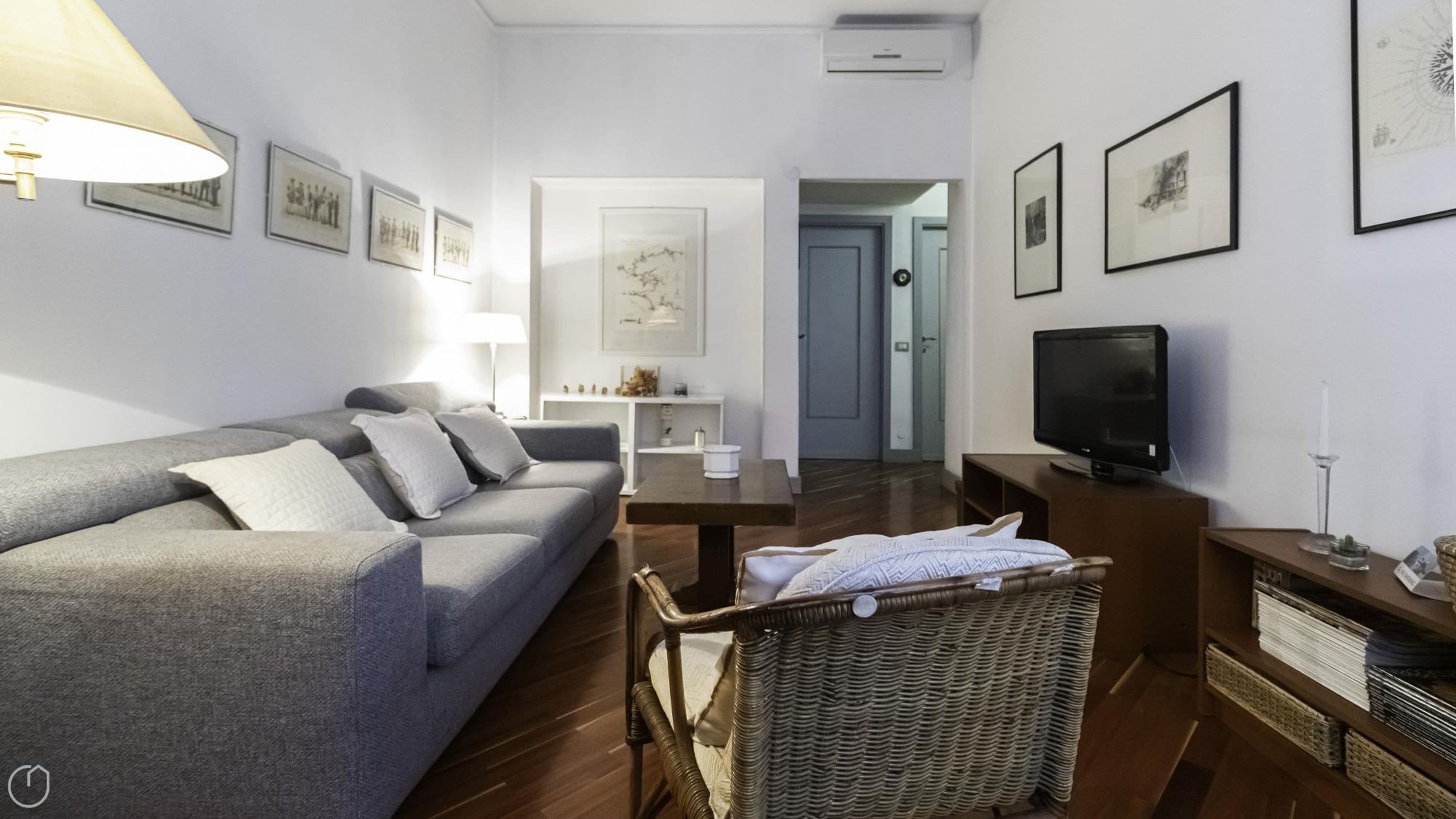 Uma sala de estar moderna com um sofá cinza, uma poltrona de vime, uma televisão e quadros nas paredes.