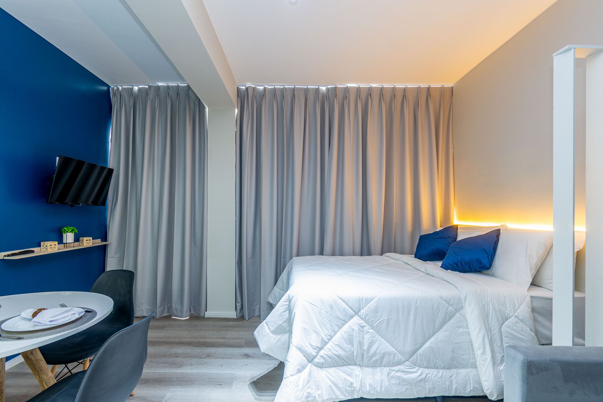 Quarto de hotel com cama, televisão na parede, mesa de jantar com cadeiras e uma pequena estante com objetos.