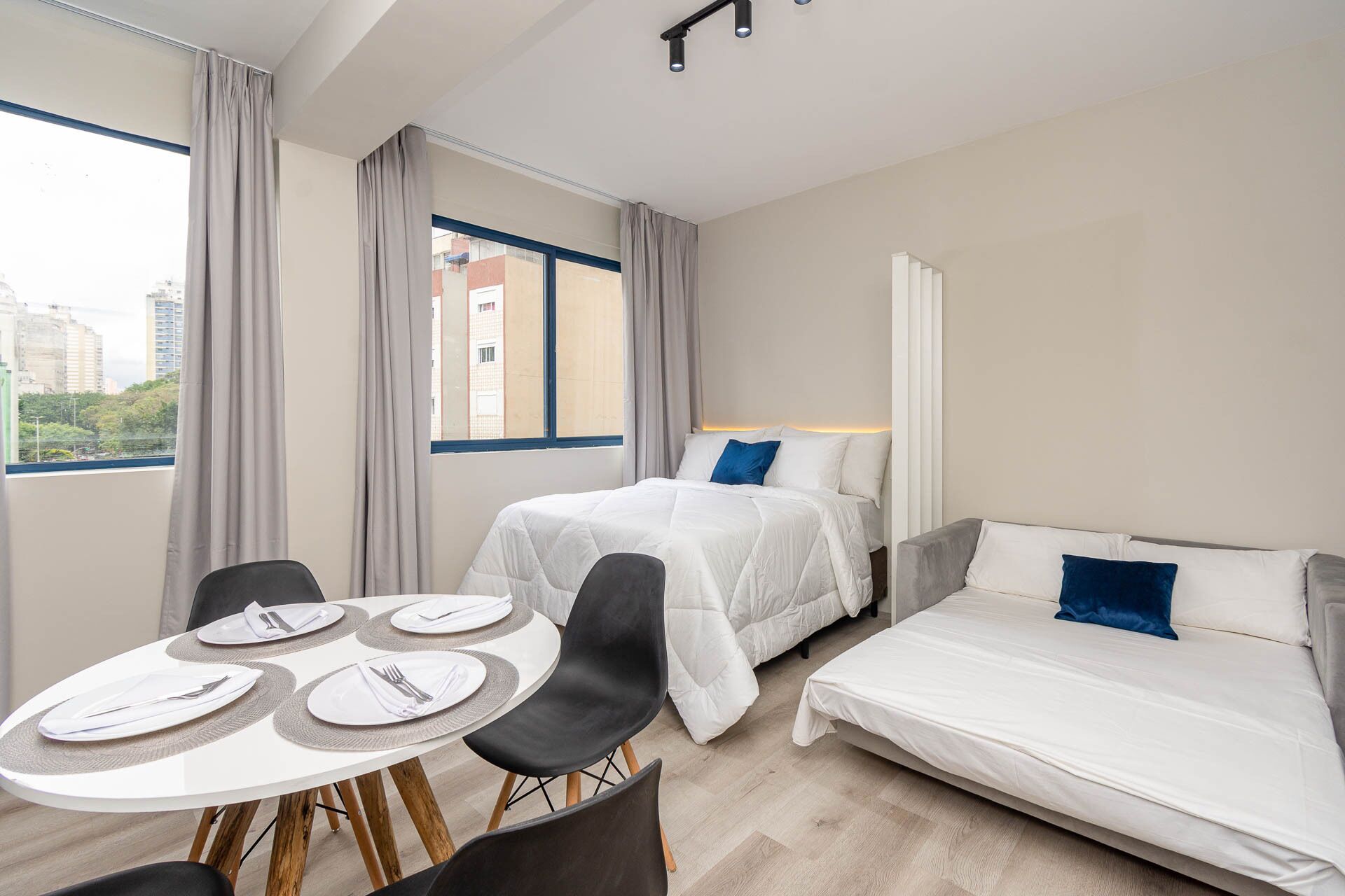 Quarto de hotel moderno com cama, sofá, mesa de jantar com cadeiras e uma grande janela com cortinas.