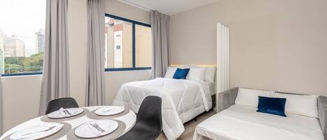 Quarto de hotel moderno com cama, sofá, mesa de jantar com cadeiras e uma grande janela com cortinas.