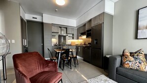 Cozinha moderna com eletrodomésticos de aço inoxidável, área de jantar com mesa e cadeiras, e sala de estar com sofá e almofadas decorativas.