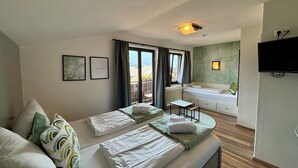 Quarto de hotel com cama, uma pequena mesa, um sofá, uma janela com cortinas, televisão na parede e banheiro com banheira.
