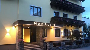 Um edifício com uma placa que diz "MAGALI'S" na entrada, com degraus de pedra, uma varanda florida e uma parede de pedra.