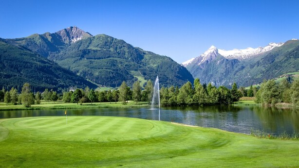 Um campo de golfe com uma fonte, rodeado por montanhas e árvores.