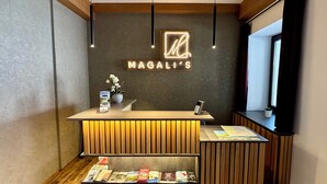 Uma receção com acabamento em madeira, um letreiro de néon com a inscrição "MAGALI'S" e uma pequena planta em vaso no balcão.