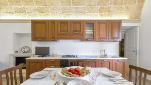 Uma área de refeições com uma mesa posta para dois, armários de madeira acima, uma lareira e uma cozinha ao fundo.