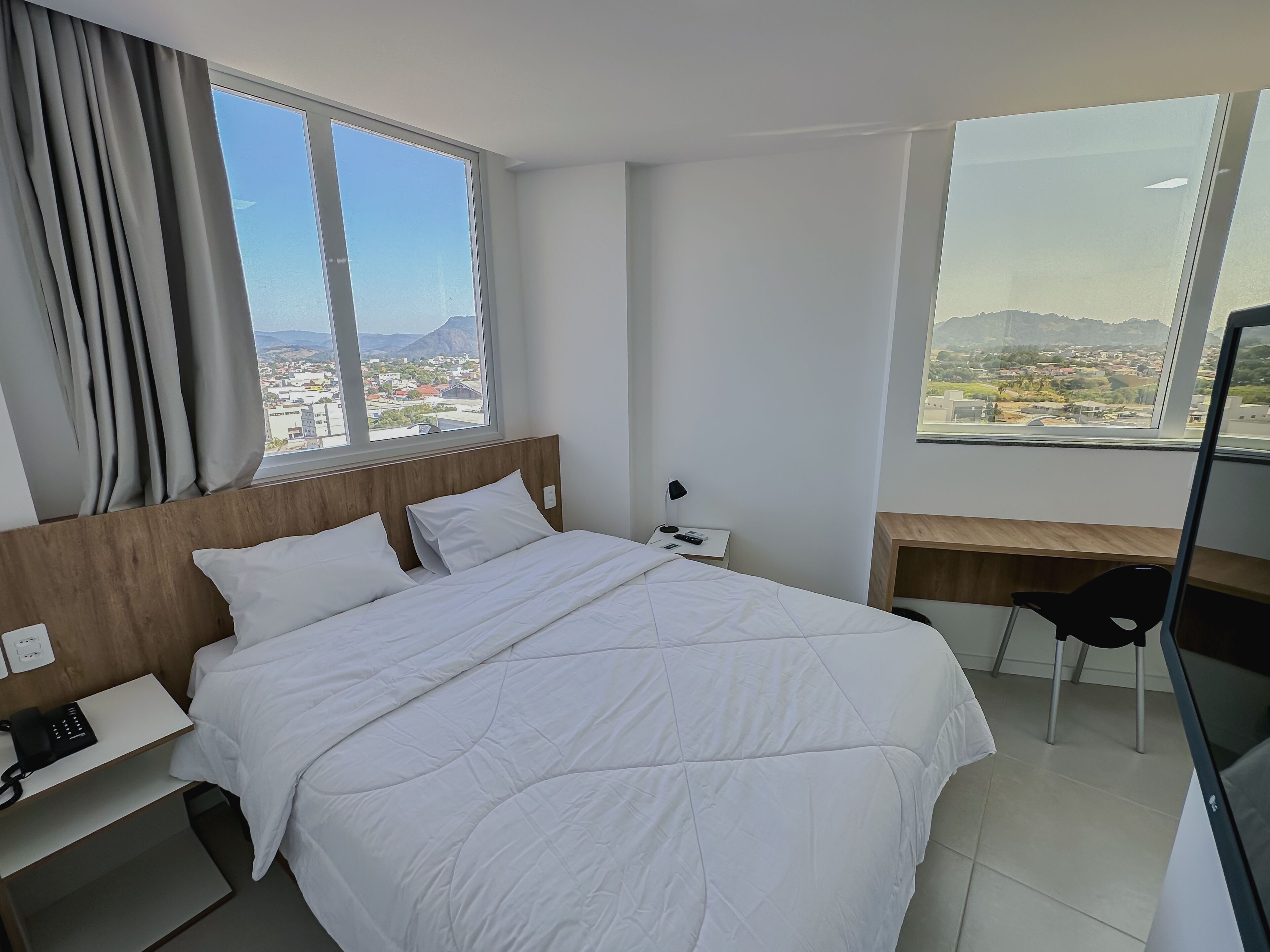 Quarto de hotel com cama, escrivaninha, cadeira, televisão e janela com vista para a cidade.
