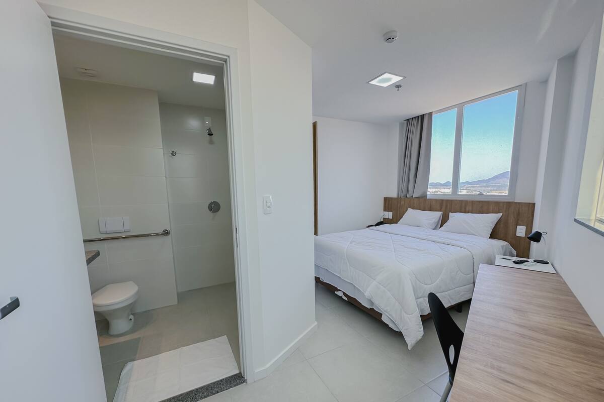 Quarto de hotel com cama, mesa de cabeceira e vista para o exterior pela janela.