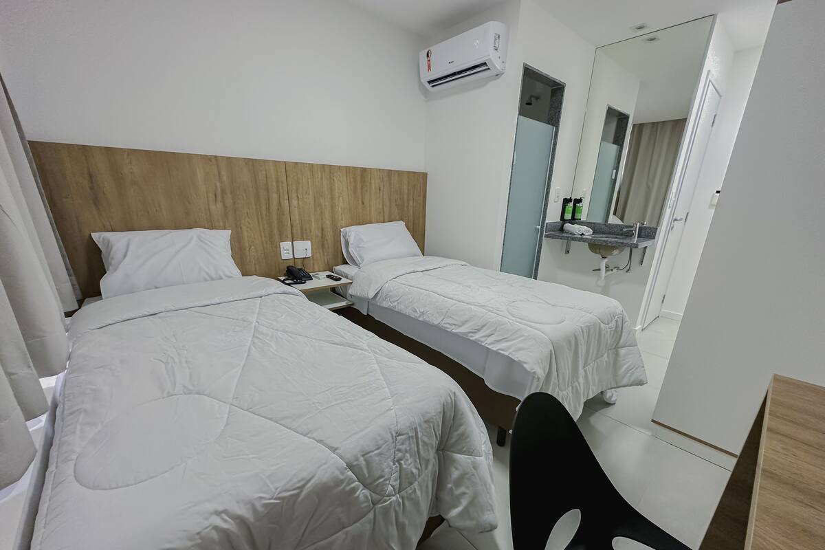 Quarto de hotel com duas camas de solteiro, cabeceira de madeira, escrivaninha com cadeira, espelho e prateleira pequena com produtos de higiene pessoal.