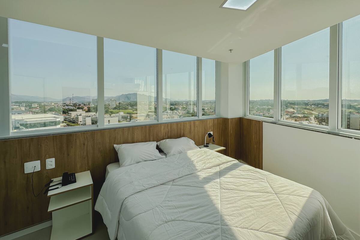 Quarto com uma cama grande, mesa de cabeceira com abajur e vista para a cidade através de amplas janelas.